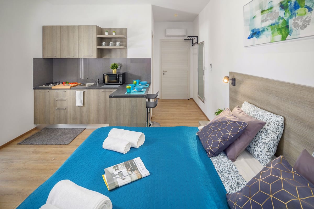 Quarto de hotel compacto com cozinha compacta, cama com roupa de cama azul e uma pequena área de refeições.