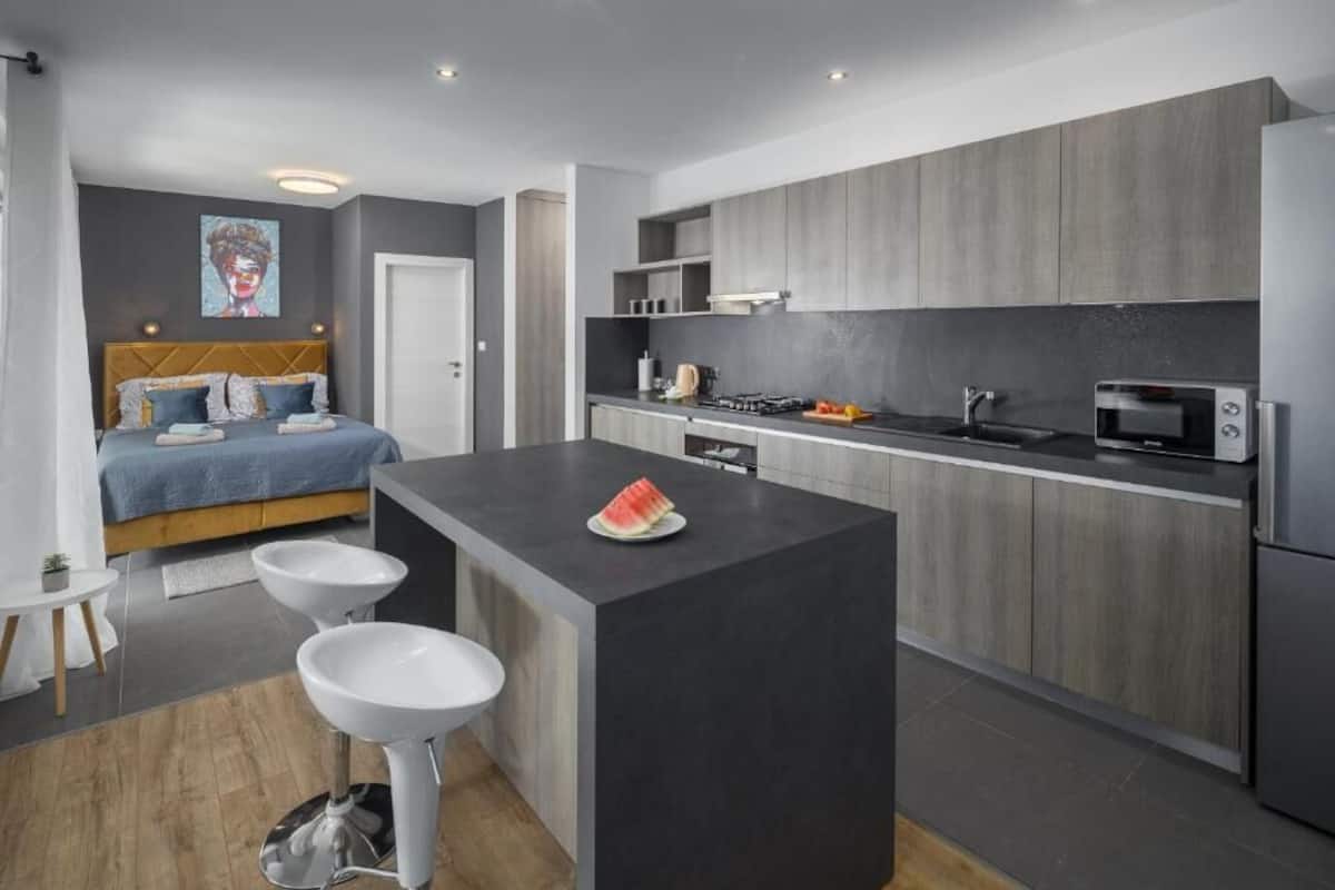 Quarto de hotel moderno com cama, mesas de cabeceira, pequena cozinha com micro-ondas e uma bancada grande com banquetas.