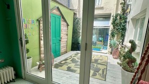 Visão através de uma porta de vidro deslizante para um pátio com porta verde, uma pequena estrutura de madeira e plantas em vasos.