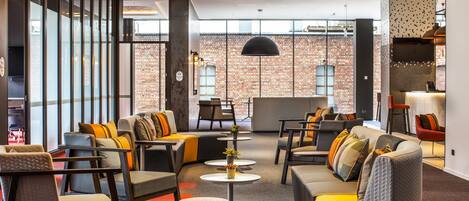 Espaço de lounge moderno com sofás, cadeiras e mesinhas, com parede de tijolos e janelas amplas.