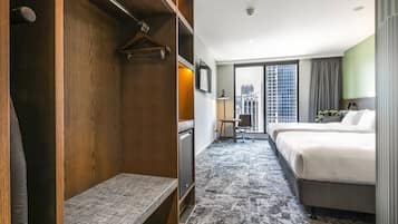 Quarto de hotel com guarda-roupa de madeira, um banco, duas camas, uma escrivaninha e uma janela grande com vista para a cidade.
