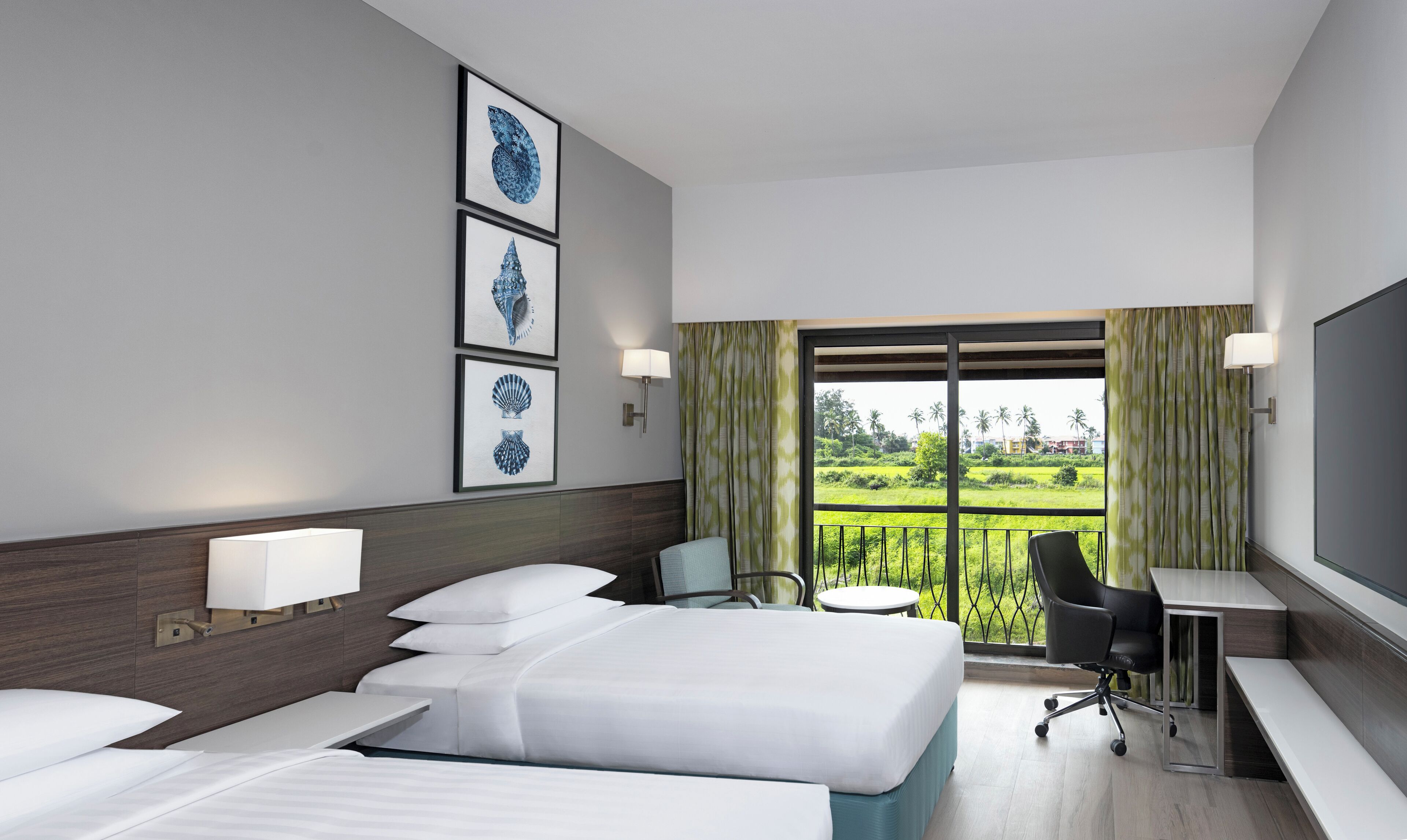Quarto de hotel com uma cama grande, uma mesa, uma cadeira e uma televisão. Há três quadros na parede e uma janela com vista para áreas verdes e edifícios.
