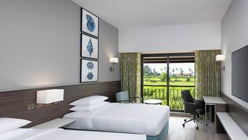 Quarto de hotel com uma cama grande, uma mesa, uma cadeira e uma televisão. Há três quadros na parede e uma janela com vista para áreas verdes e edifícios.