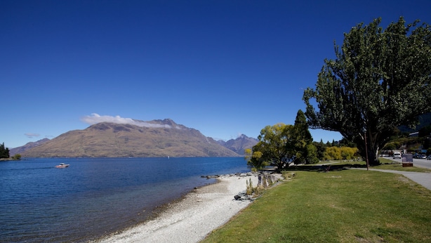 Um lago com vista clara para montanhas ao fundo, uma praia de areia e uma área gramada com árvores.