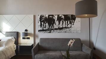 Quarto moderno com cama, sofá cinza, orquídea em vaso, luminária na parede e fotografia em preto e branco de cavalos.