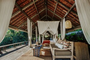 Um deck de madeira com uma cama com dossel, um banco e um lustre pendurado.