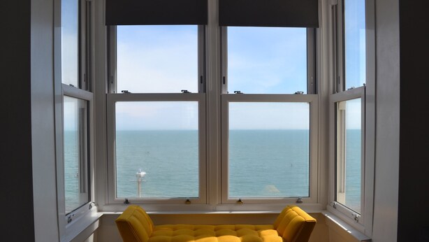 Quarto de canto com uma grande janela com vista para o mar, um banco acolchoado amarelo e um radiador branco.