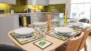 Uma cozinha moderna com armários brancos, uma área de jantar com uma mesa redonda posta para quatro pessoas, e vista para o exterior através de uma grande janela.