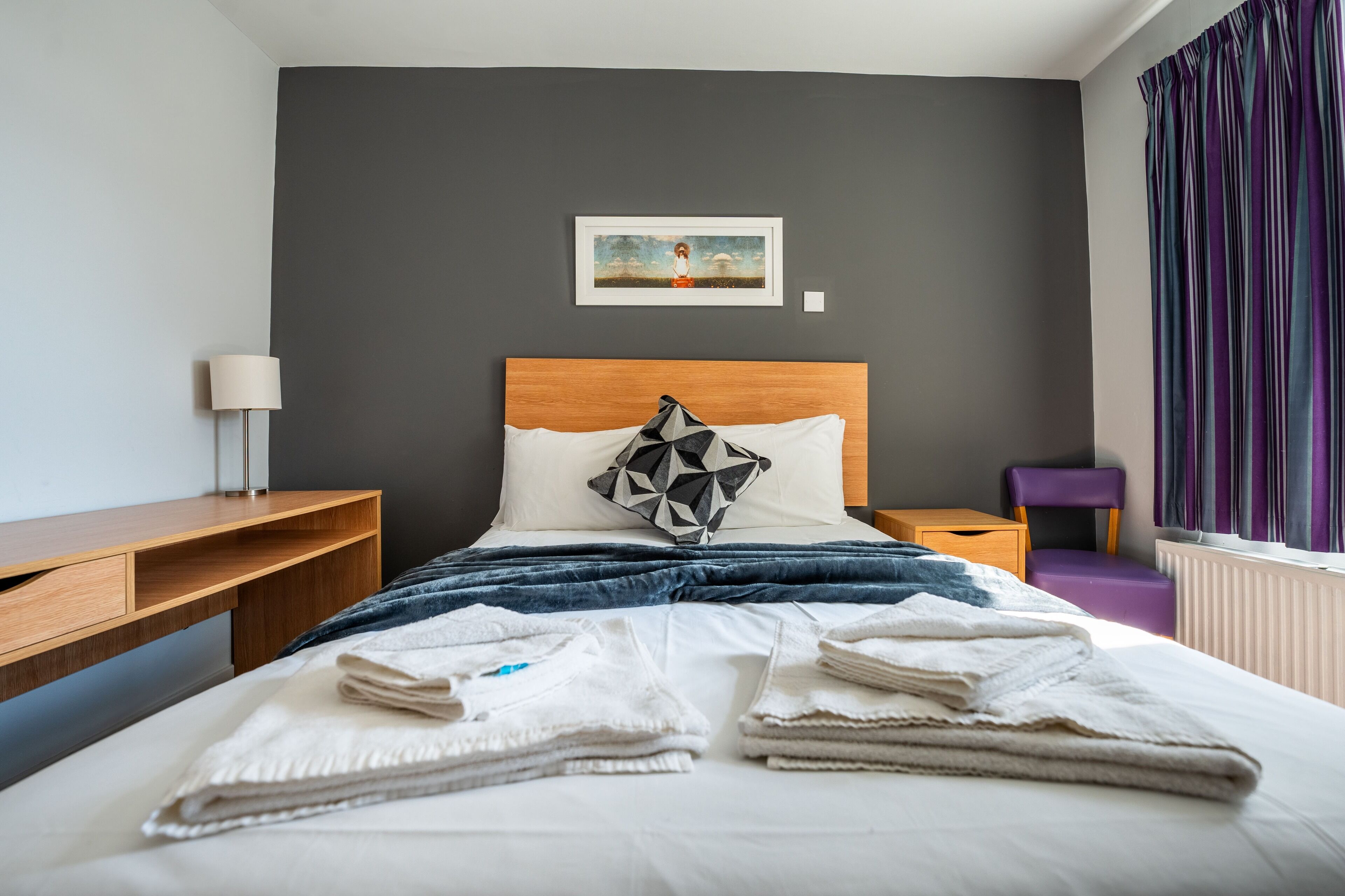Quarto de hotel com cama, mesa de cabeceira, luminária, cadeira e janela com cortinas.