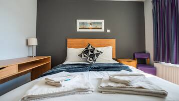 Quarto de hotel com cama, mesa de cabeceira, luminária, cadeira e janela com cortinas.