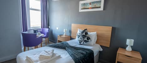Quarto de hotel com uma cama, escrivaninha, cadeira, mesa de cabeceira e um quadro na parede.