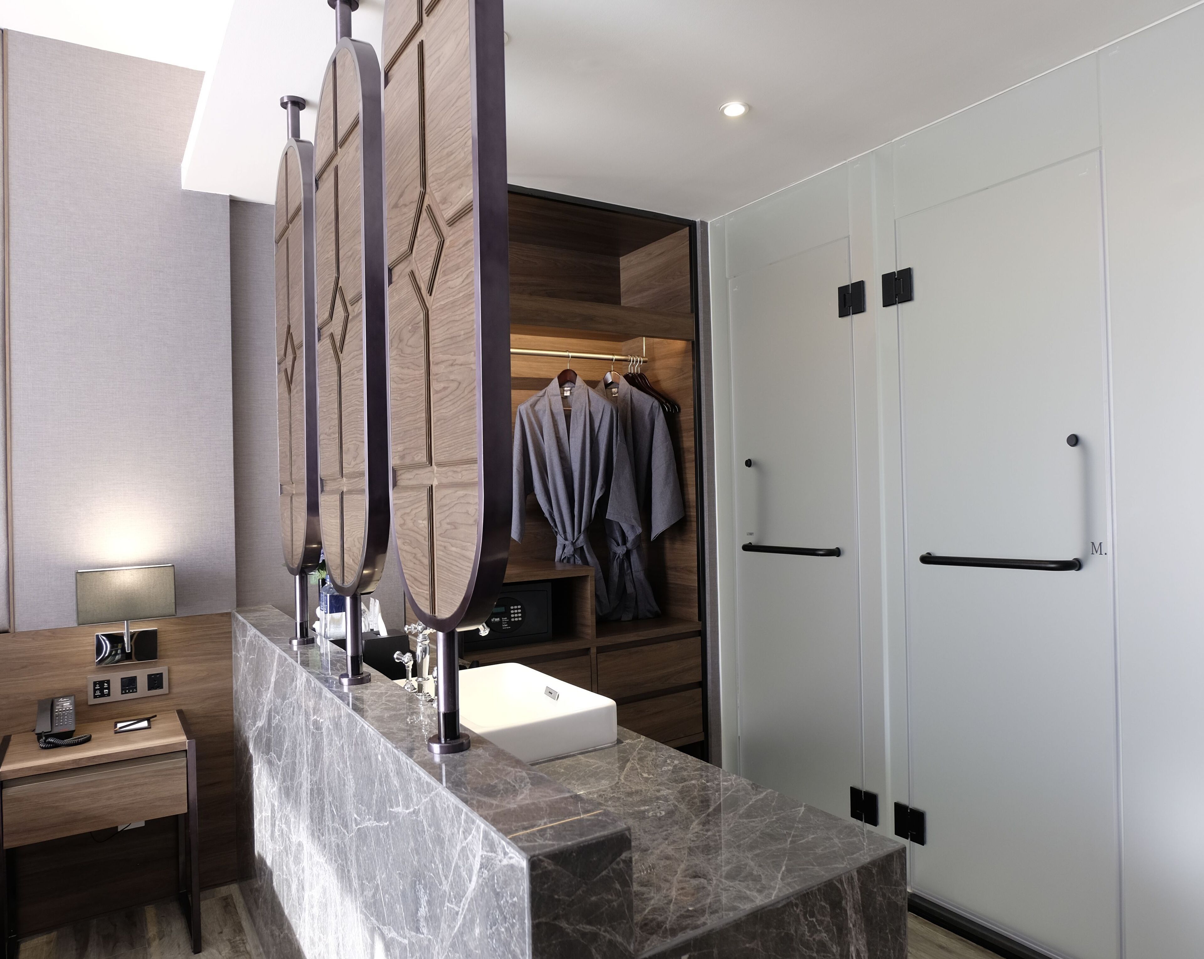 Casa de banho moderna com bancada de mármore, lavatório embutido e espelho com moldura de madeira. Apresenta uma cómoda de madeira com um telefone e uma lâmpada, e um closet espaçoso com roupões pendurados.