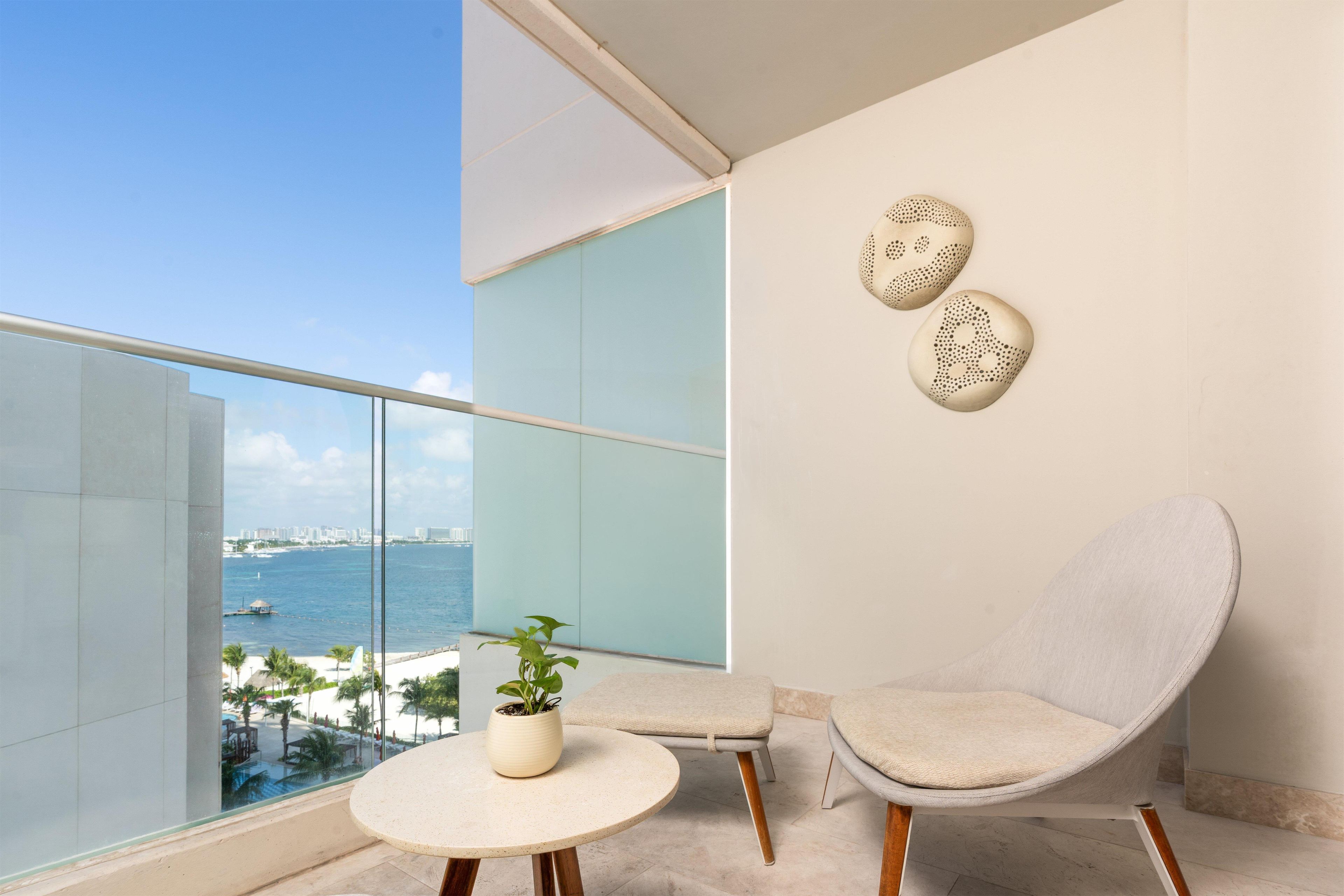 Uma varanda com vista para o mar, uma mesa redonda com uma planta, um banco acolchoado e dois objetos decorativos fixados na parede.