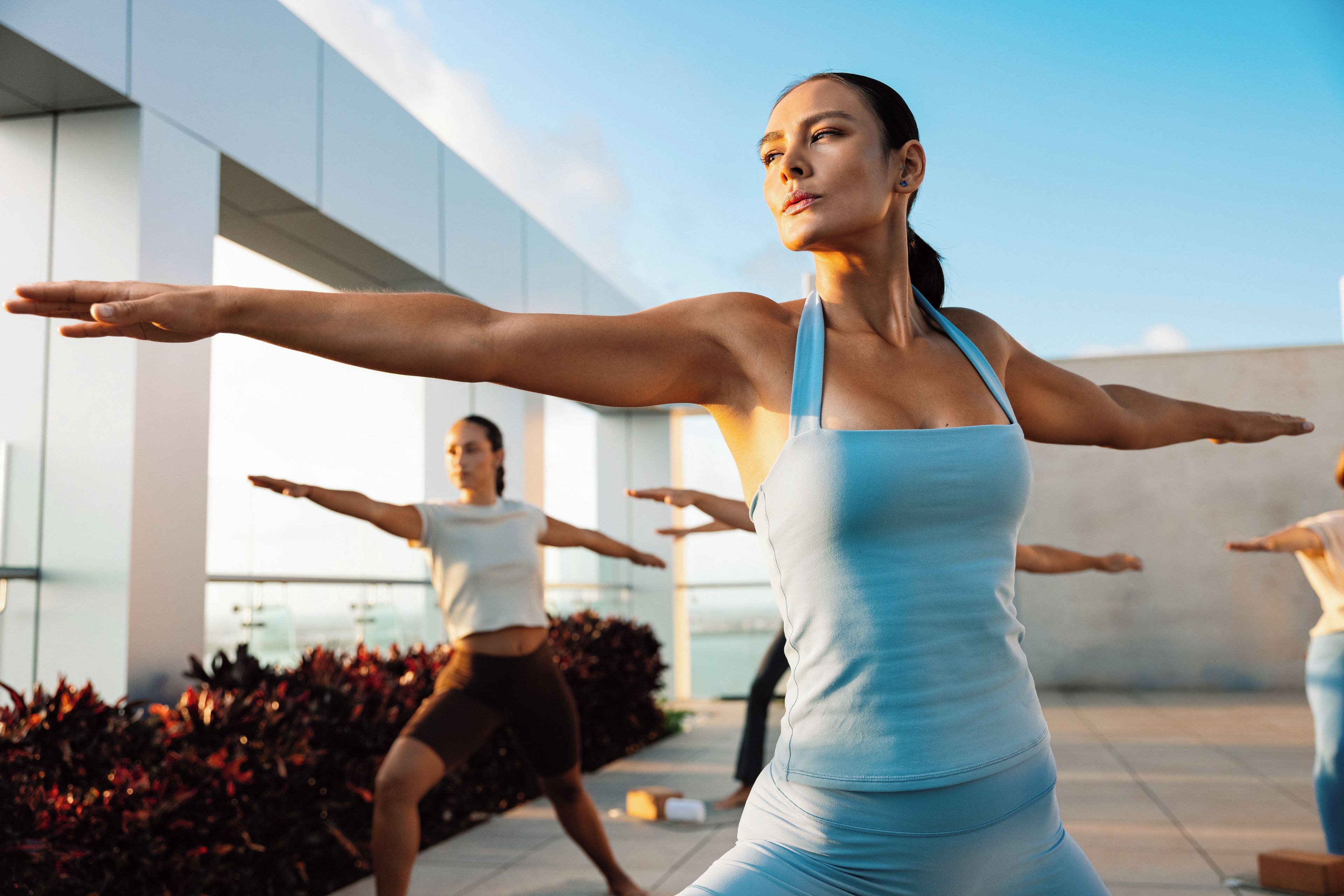 Um grupo de pessoas praticando uma postura de yoga ao ar livre, com um edifício moderno ao fundo.