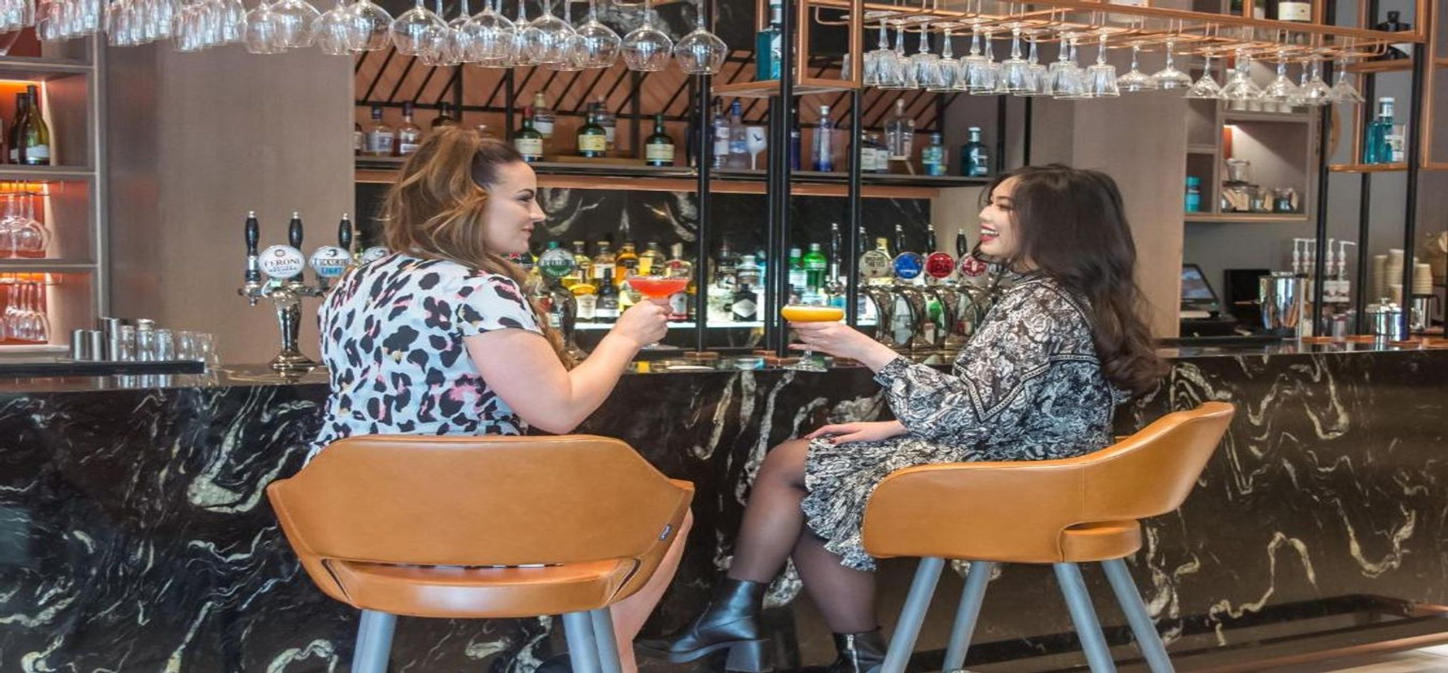 Duas pessoas sentadas num bar com bancada de mármore, rodeadas por prateleiras de garrafas e copos.