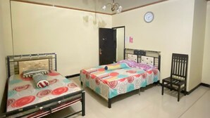 Quarto com duas camas, uma cadeira, um ventilador de teto e um relógio na parede.