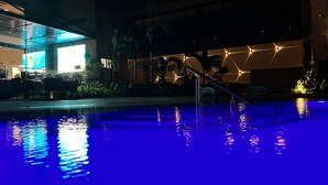 Uma área de piscina à noite, com superfície da água iluminada em azul, palmeiras e um edifício moderno ao fundo com padrões de luz geométricos.