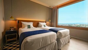 Quarto de hotel com duas camas, mesa de cabeceira, abajur e vista para a cidade.