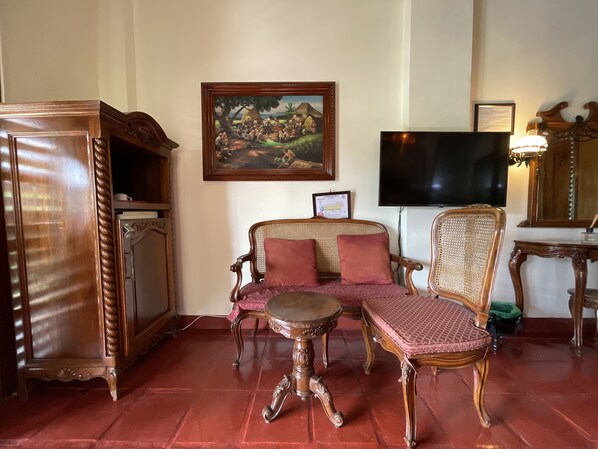Uma sala de estar tradicional com um sofá, poltrona e uma pequena mesa. Há uma televisão fixada na parede e um quadro emoldurado acima do sofá.