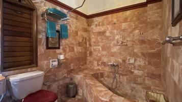 Um banheiro com parede de mármore, banheira, vaso sanitário com assento vermelho, luminária pendente e um pequeno banco com padrão floral.