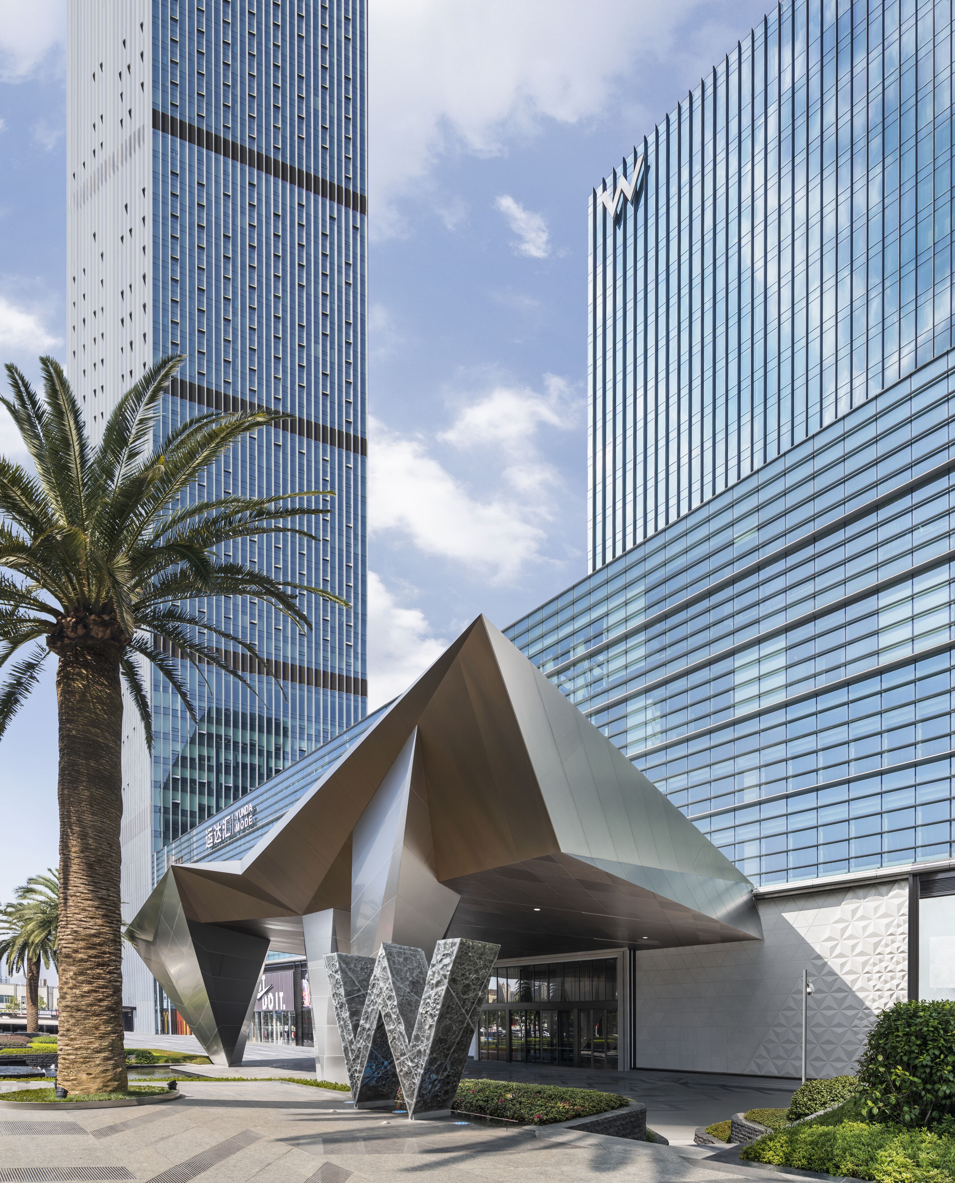 Um edifício moderno com uma estrutura de entrada geométrica marcante, rodeado por arranha-céus e palmeiras.