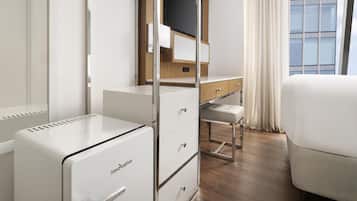 Quarto de hotel com uma kitchenette compacta, uma escrivaninha com cadeira, uma cama e uma janela grande com cortinas.