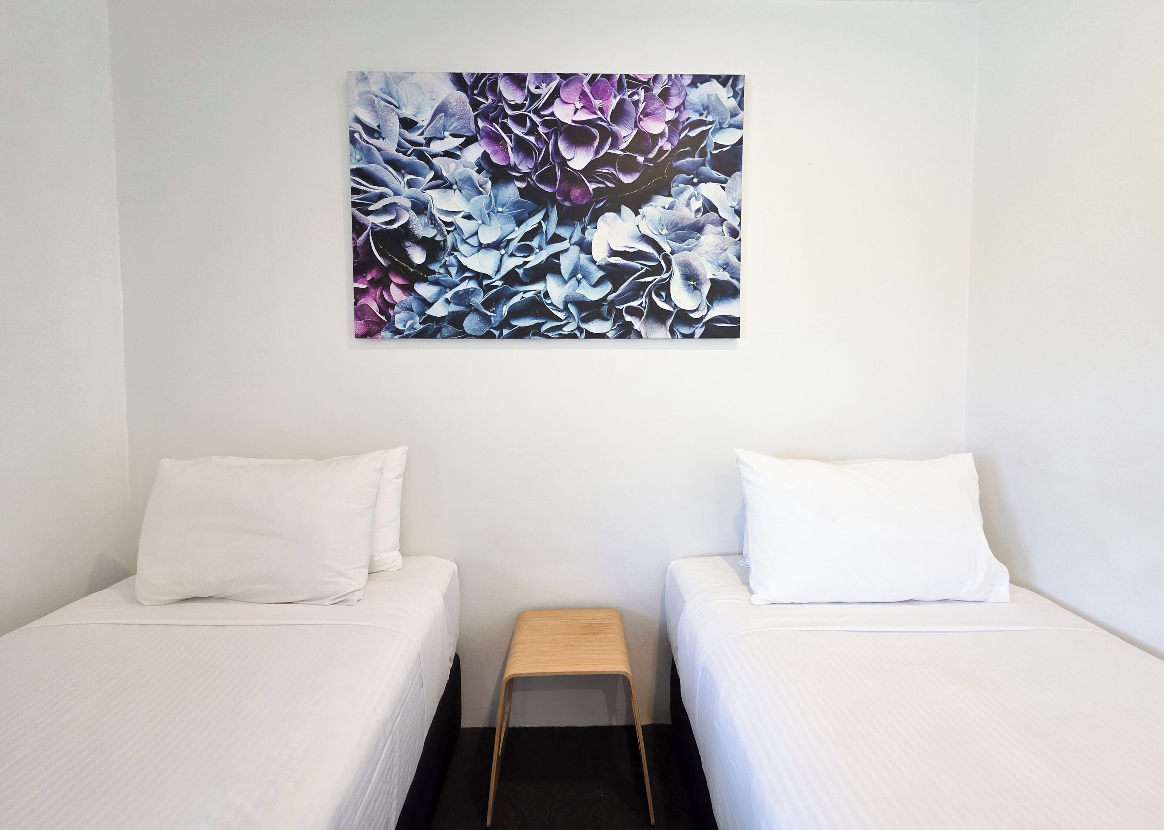 Quarto com duas camas de solteiro, um banquinho de madeira e um quadro abstrato grande na parede.