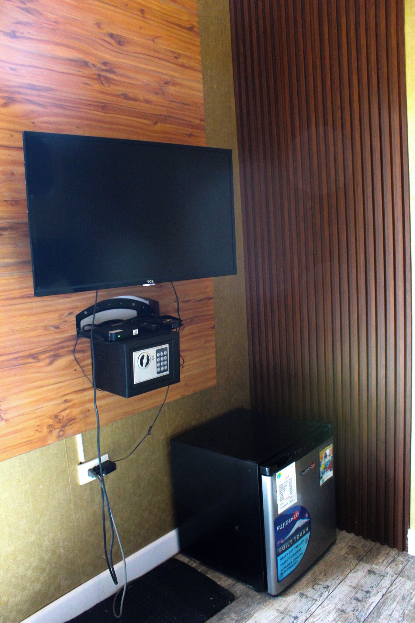 Quarto com televisão de tela plana presa à parede de madeira, dispositivo eletrônico preto em um suporte e geladeira preta com um adesivo.