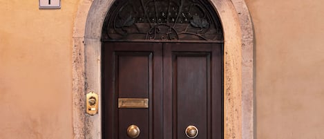 Uma porta de madeira escura com puxador e placa de latão, emoldurada por um arco de pedra, contra uma parede bege texturizada.