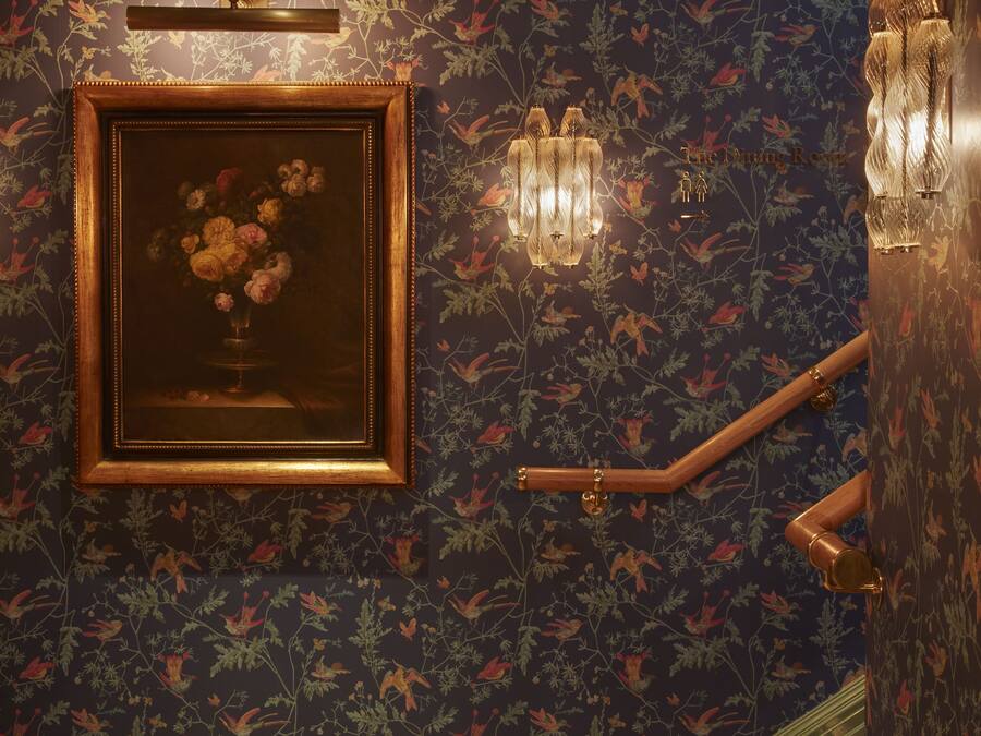 Escadaria com papel de parede floral, um quadro emoldurado de flores e luminárias fixadas na parede.