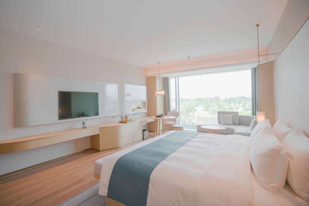 Quarto de hotel moderno com uma cama grande, televisão na parede, secretária com cadeira, mesa pequena e um sofá.