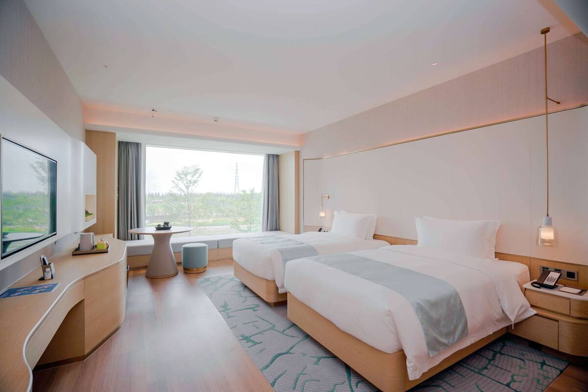 Quarto de hotel com duas camas, uma escrivaninha, uma TV e uma janela com vista para o verde.