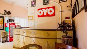 Uma área de recepção com o letreiro 'OYO', um balcão de atendimento, uma planta e um refrigerador de Coca-Cola.