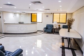 Uma área de recepção de hotel com um balcão curvo, duas poltronas azuis, uma pequena mesa com abajur e um vaso com plantas secas.
