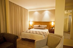 Quarto de hotel com cama, duas mesas de cabeceira, um criado-mudo, um sofá e uma cadeira.