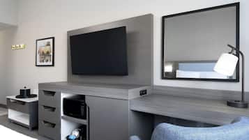 Quarto de hotel moderno com televisão de tela plana na parede, mesa de cabeceira pequena com luminária, uma cadeira e micro-ondas embutido.
