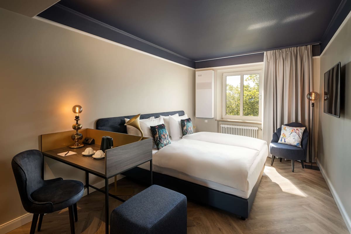 Quarto de hotel moderno com uma cama grande, uma escrivaninha, uma cadeira e uma mesinha.