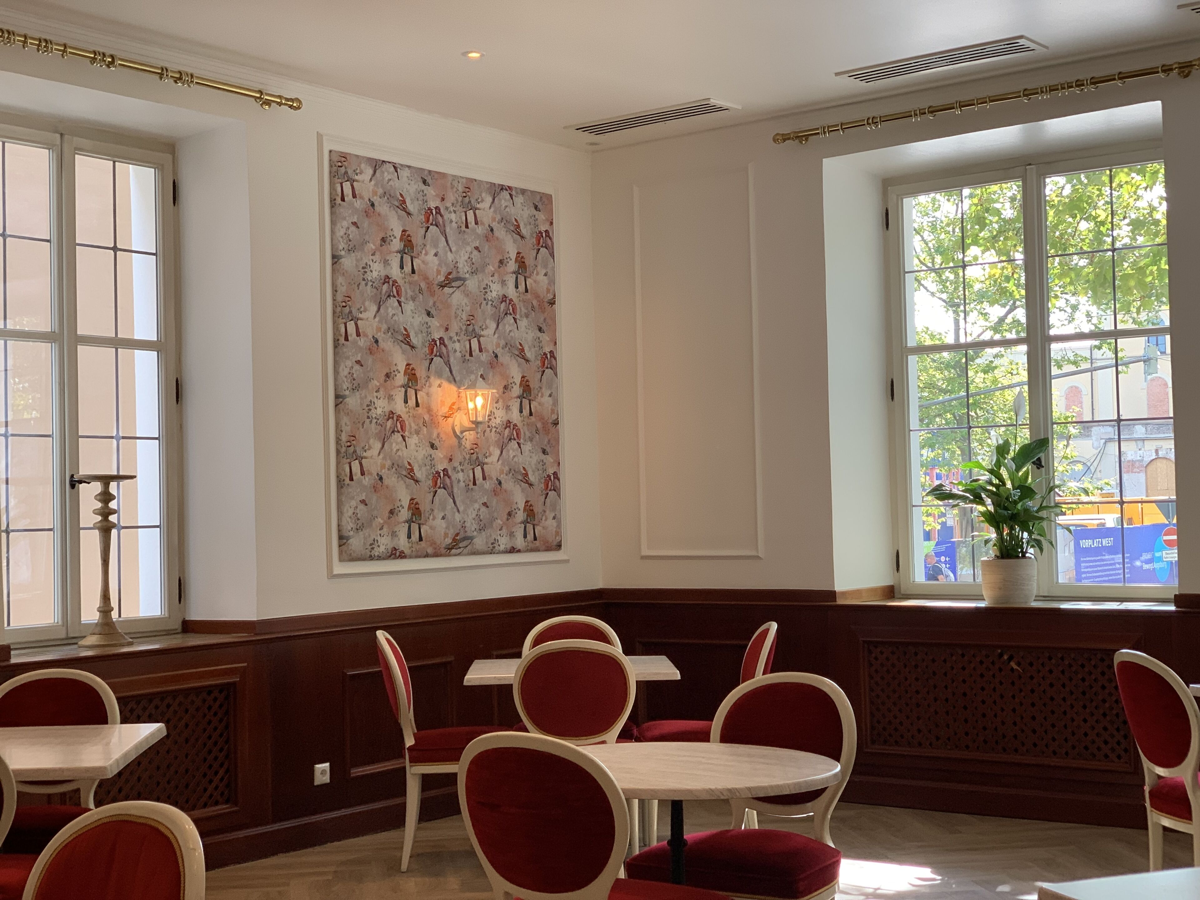 Interior de restaurante com uma grande pintura abstrata, várias mesas e cadeiras estofadas em vermelho, e vista para a rua.