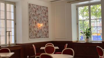 Interior de restaurante com uma grande pintura abstrata, várias mesas e cadeiras estofadas em vermelho, e vista para a rua.