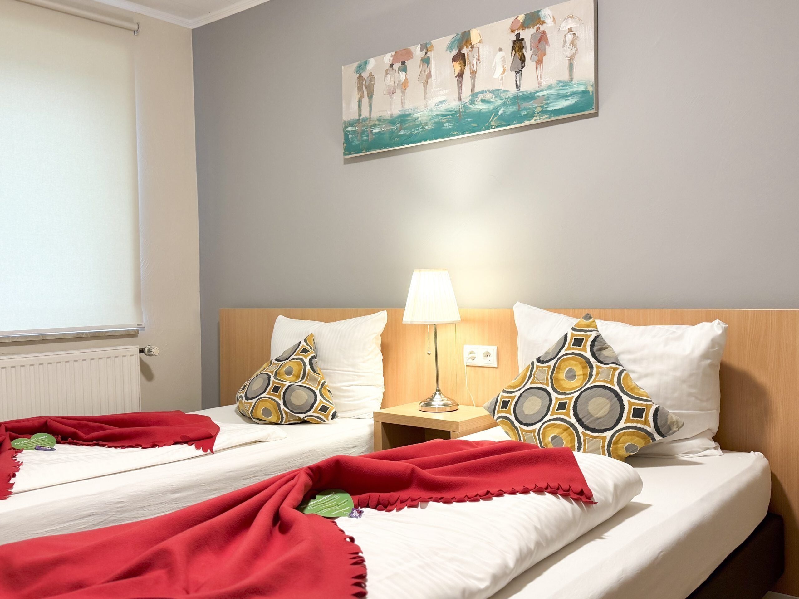 Quarto de hotel com duas camas, um quadro na parede, uma mesa de cabeceira com abajur e uma janela com persianas.