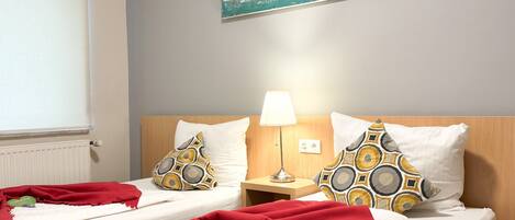 Quarto de hotel com duas camas, um quadro na parede, uma mesa de cabeceira com abajur e uma janela com persianas.