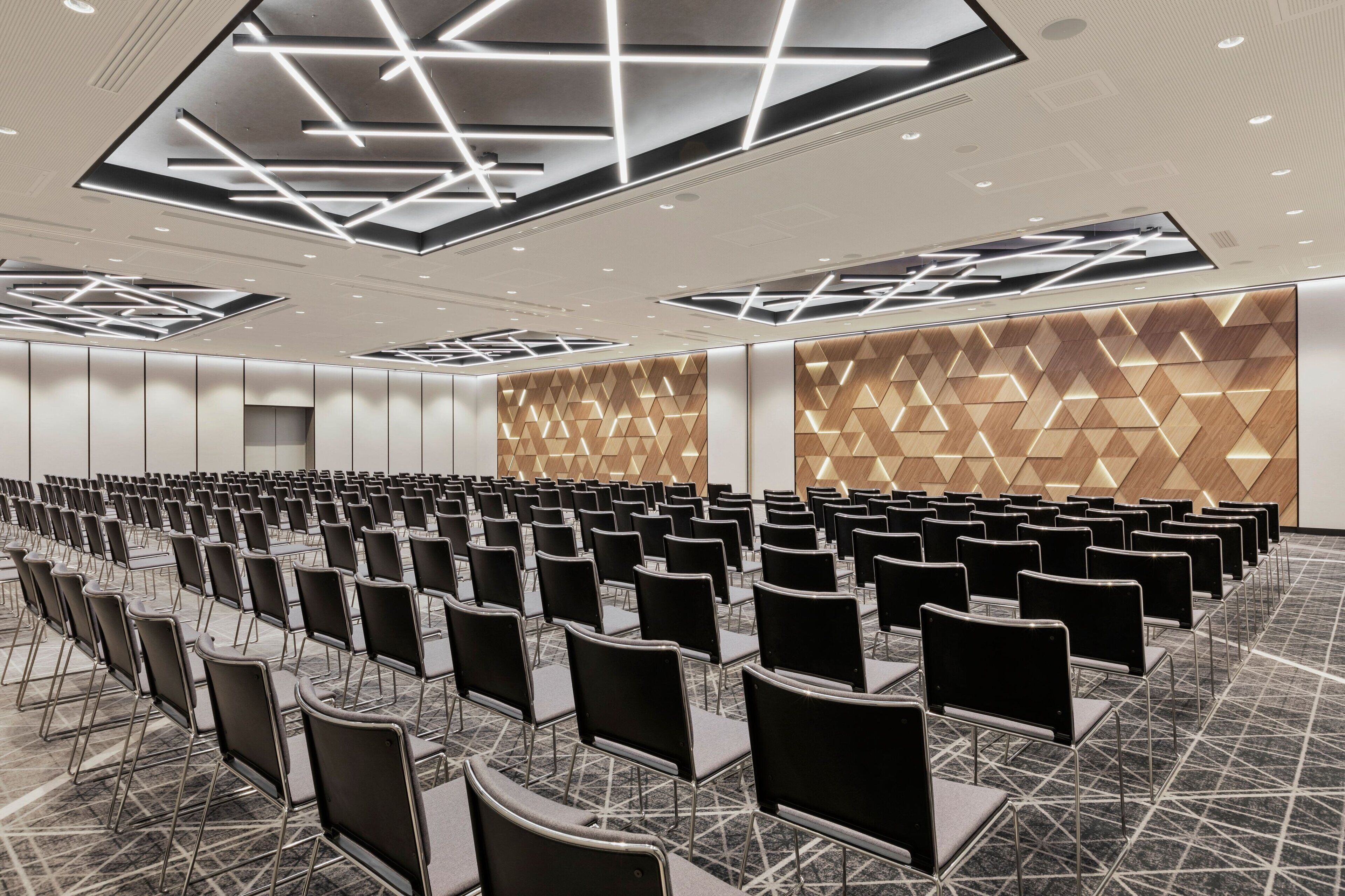 Uma sala de conferências com fileiras de cadeiras, parede estampada e luminárias de teto geométricas.