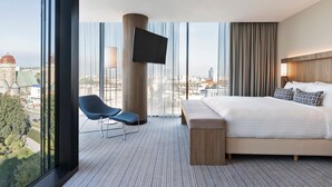 Quarto de hotel moderno com uma cama grande, uma TV de tela plana fixada em um suporte, uma cadeira azul e vista para a cidade através de grandes janelas.