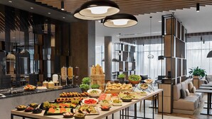 Uma área de refeições moderna com uma mesa comprida posta com diversos alimentos, um espaço de bar com balcão e uma área de estar com sofás e mesas.