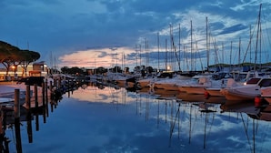 Uma marina com vários barcos atracados, água calma refletindo os barcos e o céu, e edifícios ao fundo.