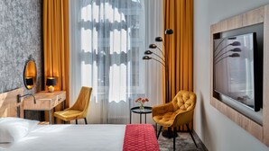 Quarto de hotel com cama, escrivaninha com luminária, cadeira, televisão e janela com cortinas.