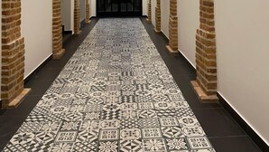 Um corredor com arcos de tijolo e ladrilhos estampados, que leva a uma entrada com porta de vidro.