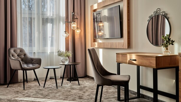 Quarto de hotel moderno com uma janela grande, televisão de tela plana na parede, escrivaninha de madeira, duas cadeiras e uma mesa pequena com um vaso de flores.