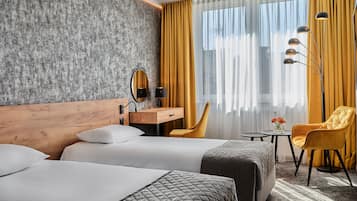 Quarto de hotel com duas camas, cabeceira de madeira, escrivaninha com espelho, uma cadeira e uma mesinha com um vaso.