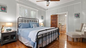 Quarto com estrutura de cama em metal, piso de madeira, ventilador de teto e área de jantar visível por uma porta aberta.